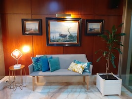 Quarto com parede de madeira, sofá cinza com almofadas estampadas, mesa pequena com abajur e planta em vaso.