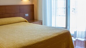 Quarto de hotel com uma cama grande, mesa de cabeceira, luminária na parede e vista pela janela com cortinas.