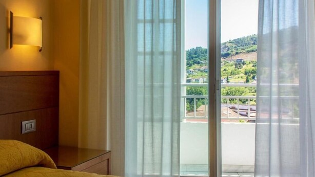 Quarto de hotel com uma cama, mesa de cabeceira, luminária, janela com cortinas e vista para a paisagem.