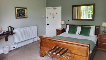 Quarto com cama de madeira, mesas de cabeceira, radiador, porta e janela com vista para o verde.