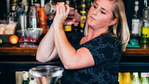 Um bartender prepara uma bebida num bar, com prateleiras cheias de garrafas de bebidas ao fundo.
