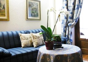 Sala de estar com sofá, mesa com toalha estampada, planta em vaso e janela com cortinas.