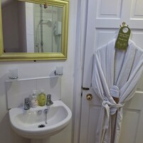 Casa de banho com uma pia branca, um espelho com moldura dourada e um roupão pendurado na porta, com uma placa que diz 'Por favor, limpe o quarto, obrigado'.