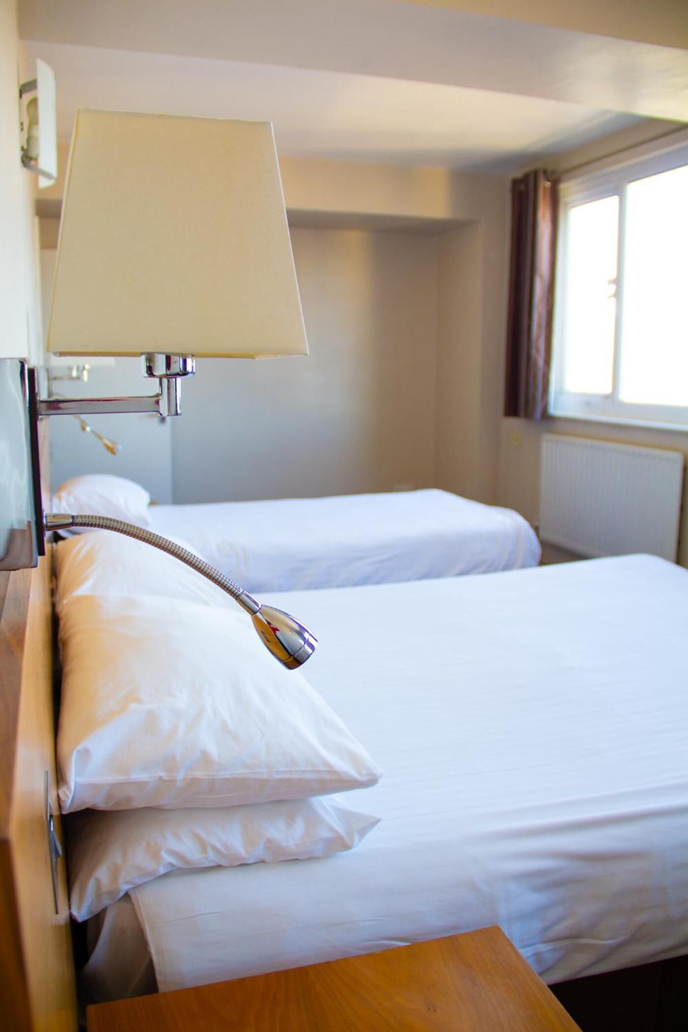 Quarto de hotel com duas camas de solteiro, uma mesa de cabeceira, uma luminária e uma janela com cortinas.