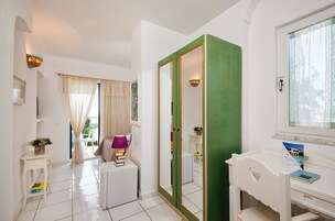 Um quarto com escrivaninha branca, espelho com moldura verde, janela com cortinas leves, mesa pequena com abajur, e porta que dá para outro cômodo.