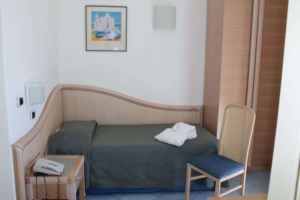 Uma cama de solteiro com colcha verde estampada, uma toalha branca na cama, uma mesinha de cabeceira com um telefone e uma cadeira de vime.