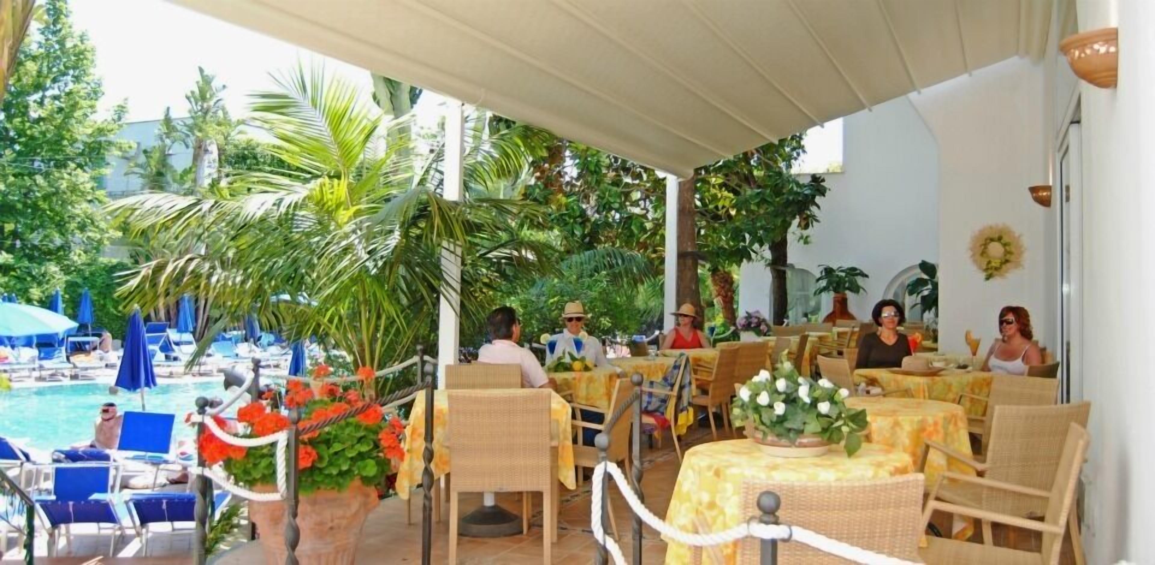 Uma área de refeições ao ar livre com cadeiras e mesas de vime, piscina e plantas tropicais.