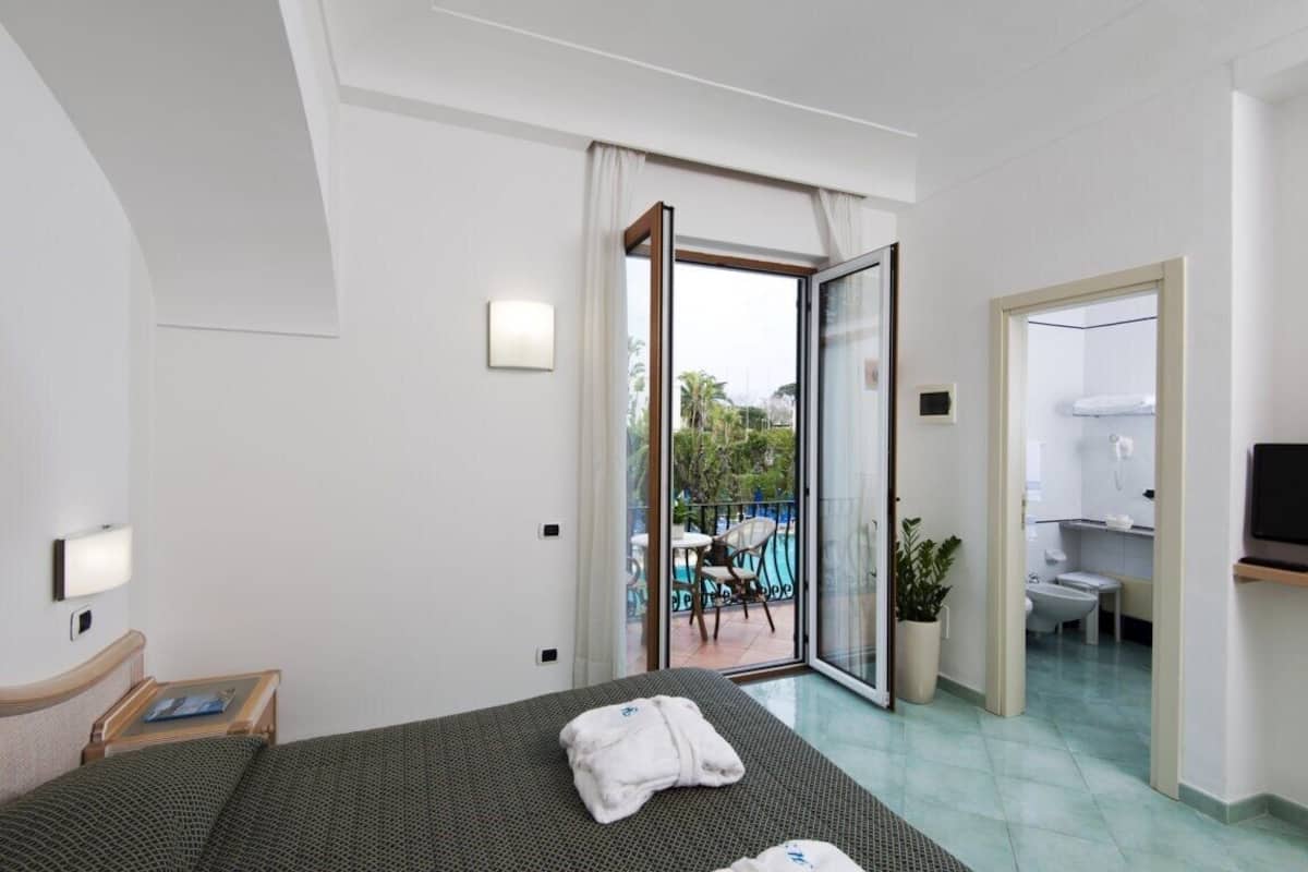 Quarto de hotel com cama, mesa pequena, televisão, banheiro e varanda com piscina.