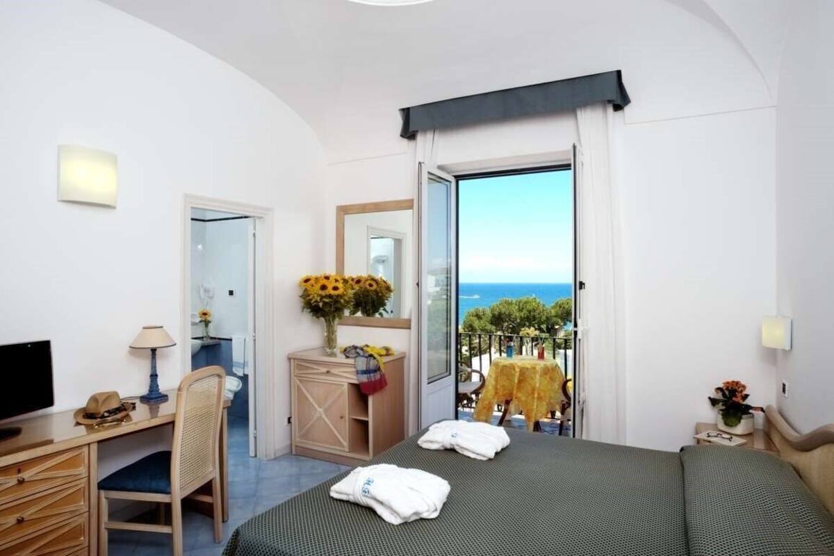 Quarto de hotel com cama, escrivaninha, cadeira, televisão, janela vista para o mar e varanda com mesa e cadeiras.
