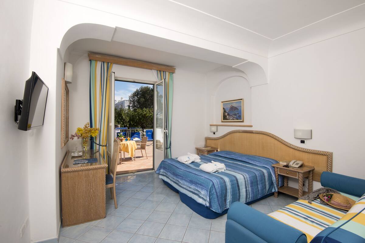 Quarto de hotel com duas camas, uma escrivaninha, uma cadeira, uma televisão e varanda com mesa e cadeiras.