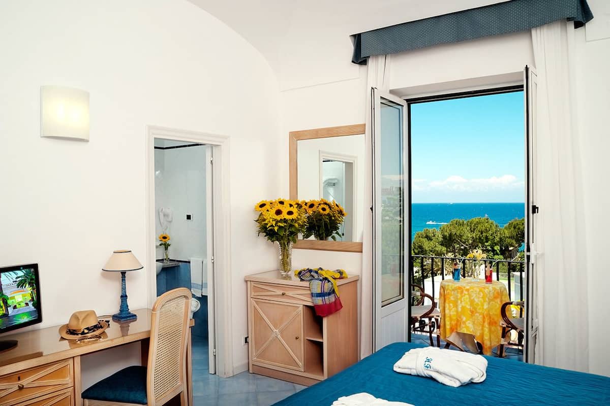Quarto de hotel com cama, secretária com computador, cadeira, cómoda e espelho, e varanda com vista para o mar.