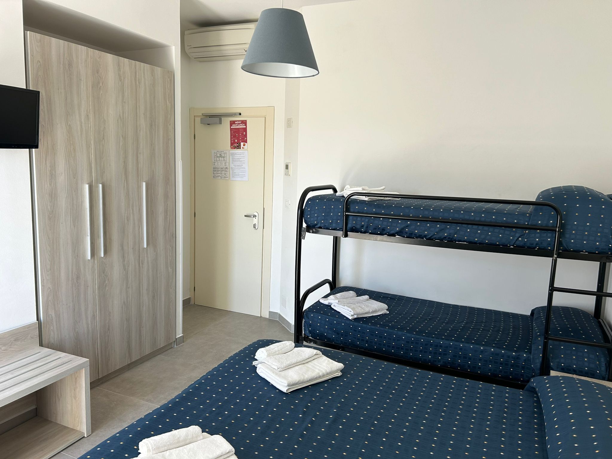Uma cama beliche com colcha estampada azul, um guarda-roupa de madeira e uma televisão à esquerda.