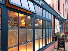 Um restaurante com fachada escura e grandes janelas, com uma placa indicando que serve comida, e um letreiro 'HOTEL' acima.