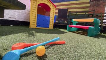 Uma área de recreação infantil com uma casinha, escorrega e diversos brinquedos sobre grama sintética.
