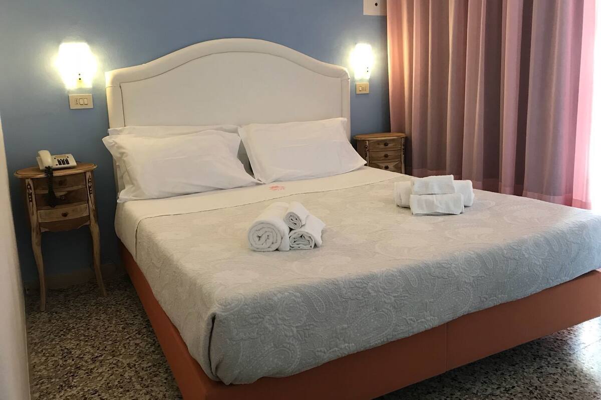 Cama bem arrumada com lençóis brancos, duas mesas de cabeceira com luminárias e uma cortina na janela.