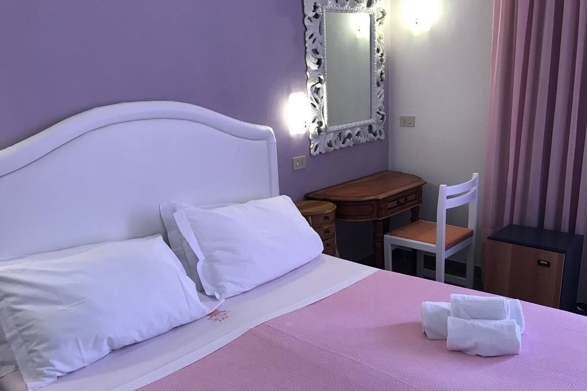 Uma cama bem arrumada com roupa de cama branca, um criado-mudo de madeira, uma cadeira e um espelho com moldura decorativa.