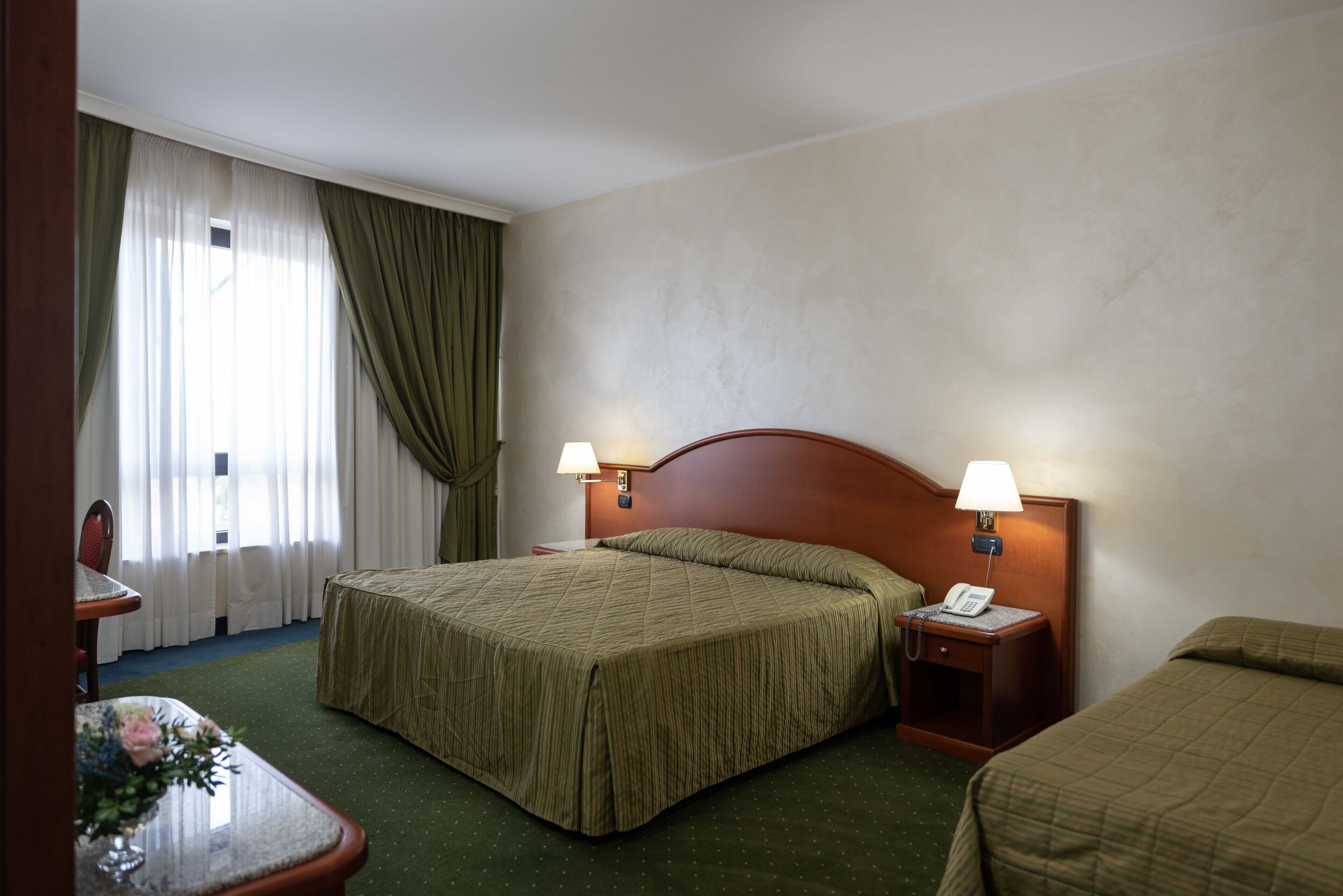 Quarto de hotel com uma cama, duas mesas de cabeceira com luminárias, uma cadeira e uma escrivaninha com um vaso de flores.