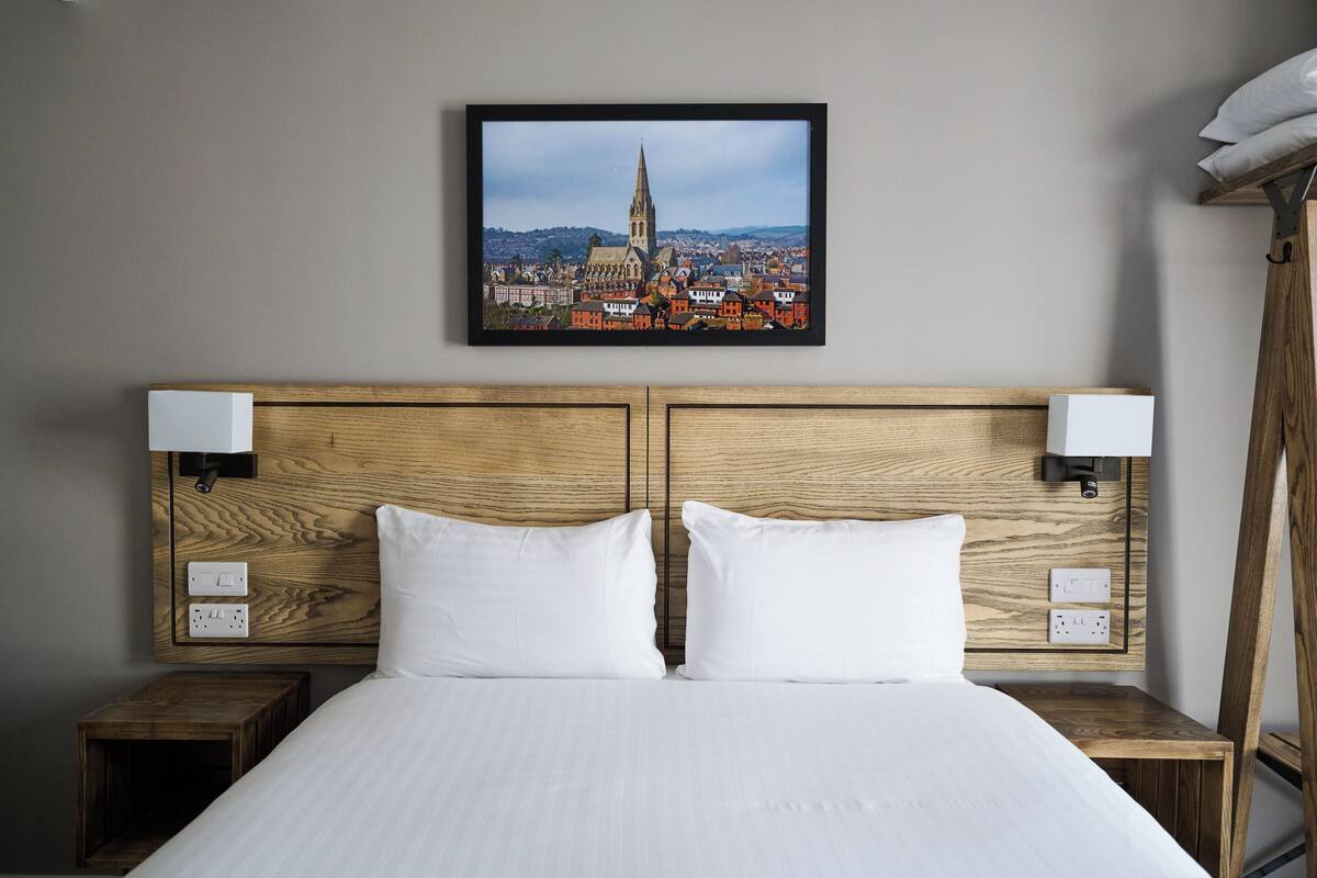 Quarto de hotel com uma cama grande, duas mesas de cabeceira, uma fotografia emoldurada da paisagem urbana na parede e um painel de cabeceira de madeira.