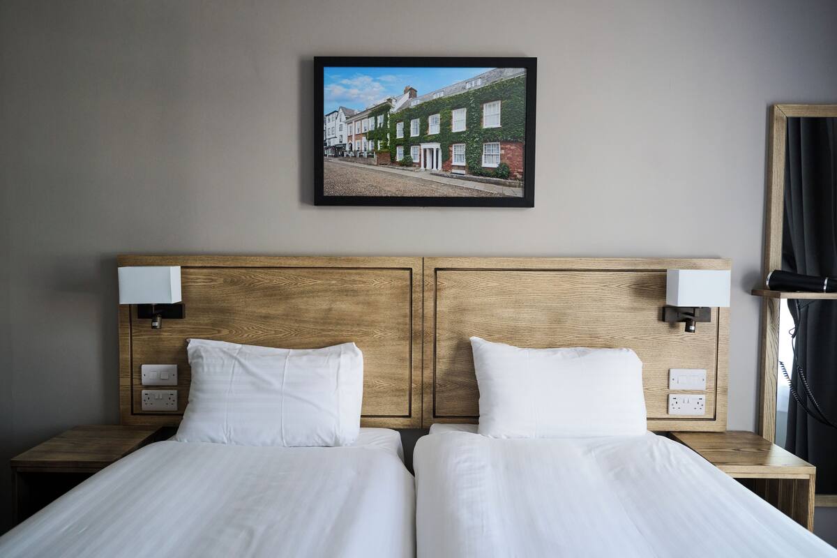 Quarto de hotel com duas camas, cabeceira de madeira, roupa de cama branca e um quadro na parede retratando uma cena de rua.
