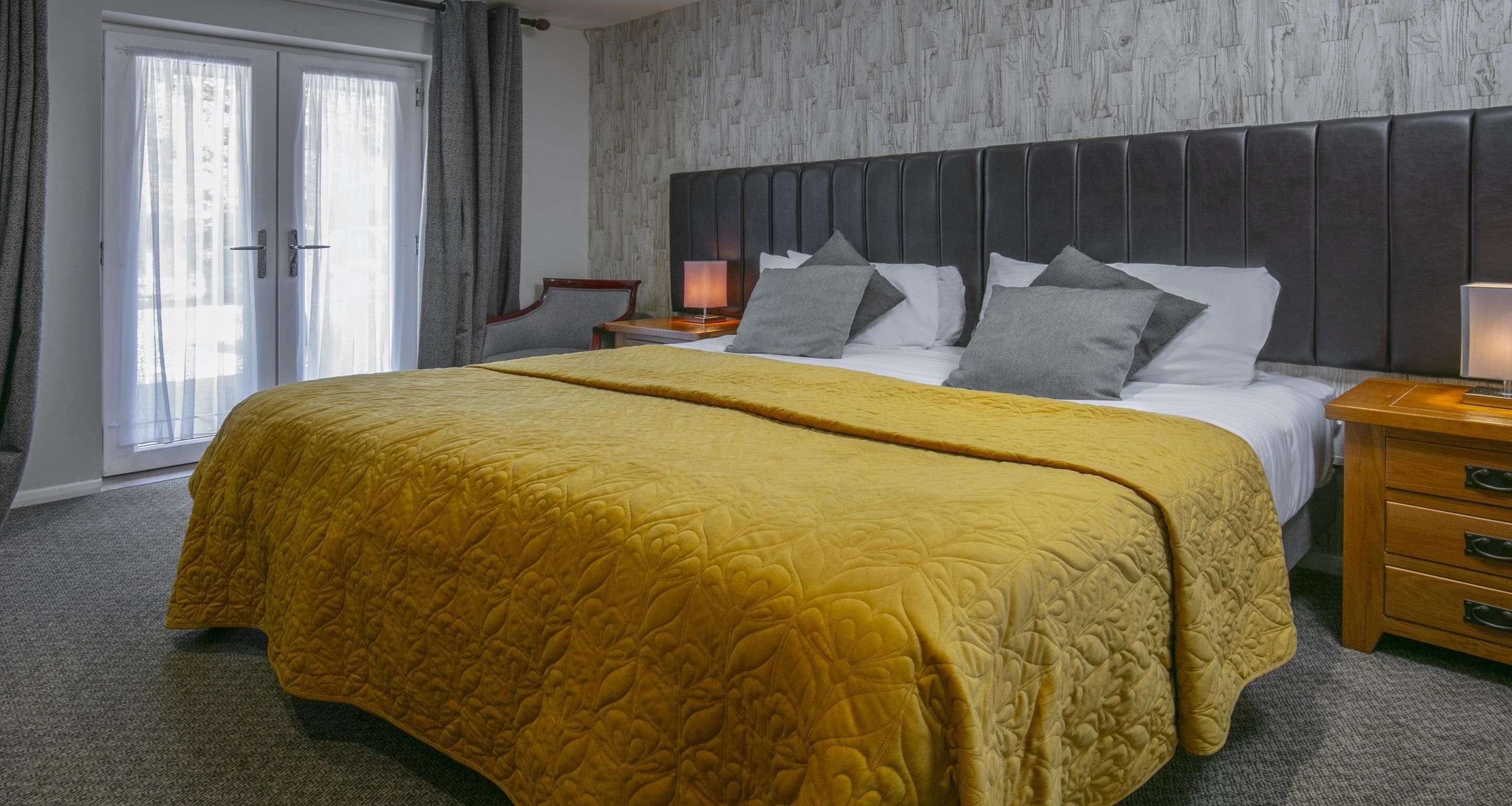 Quarto de hotel com uma cama grande, mesa de cabeceira de madeira, luminária e vista para o exterior através de uma porta com cortinas.