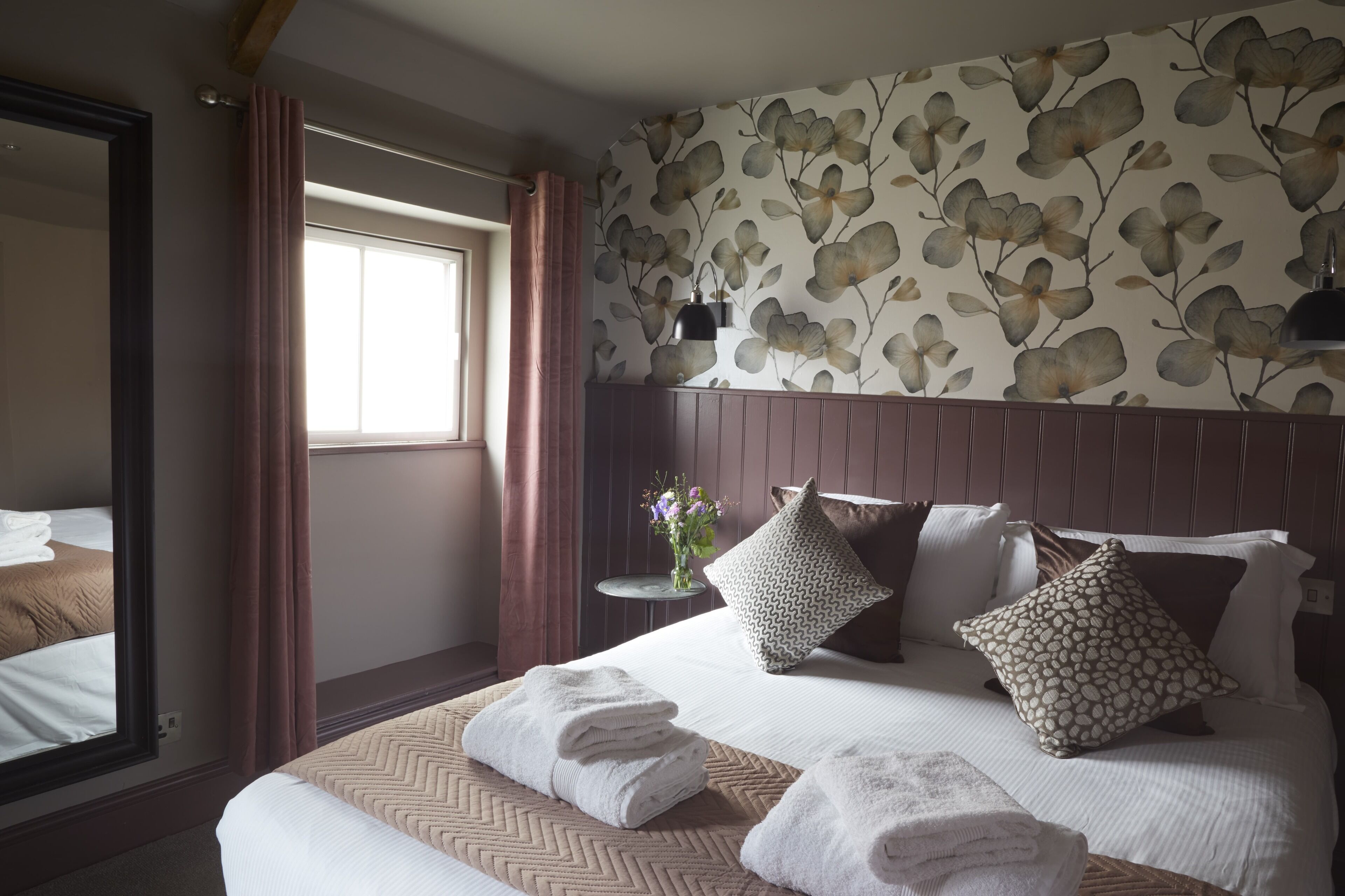Quarto de hotel com uma cama grande, um espelho, uma janela com cortinas, uma mesinha de cabeceira com um vaso de flores e uma parede com padrão floral.