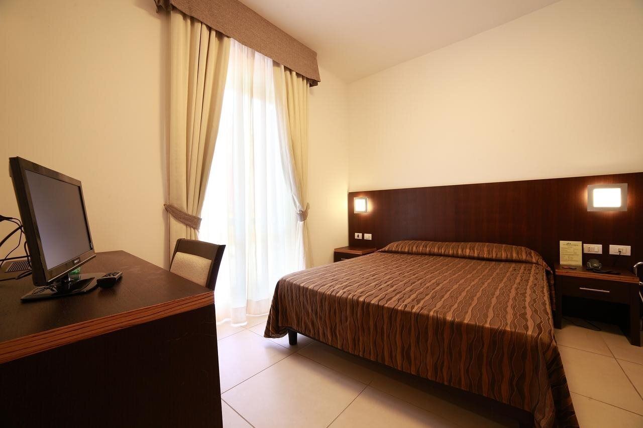 Quarto de hotel com uma cama, uma escrivaninha com computador, uma cadeira, mesa de cabeceira e janela com cortinas.