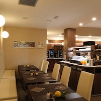 Restaurante com várias mesas, cadeiras e área de cozinha com máquinas de café e exposição de frutas.