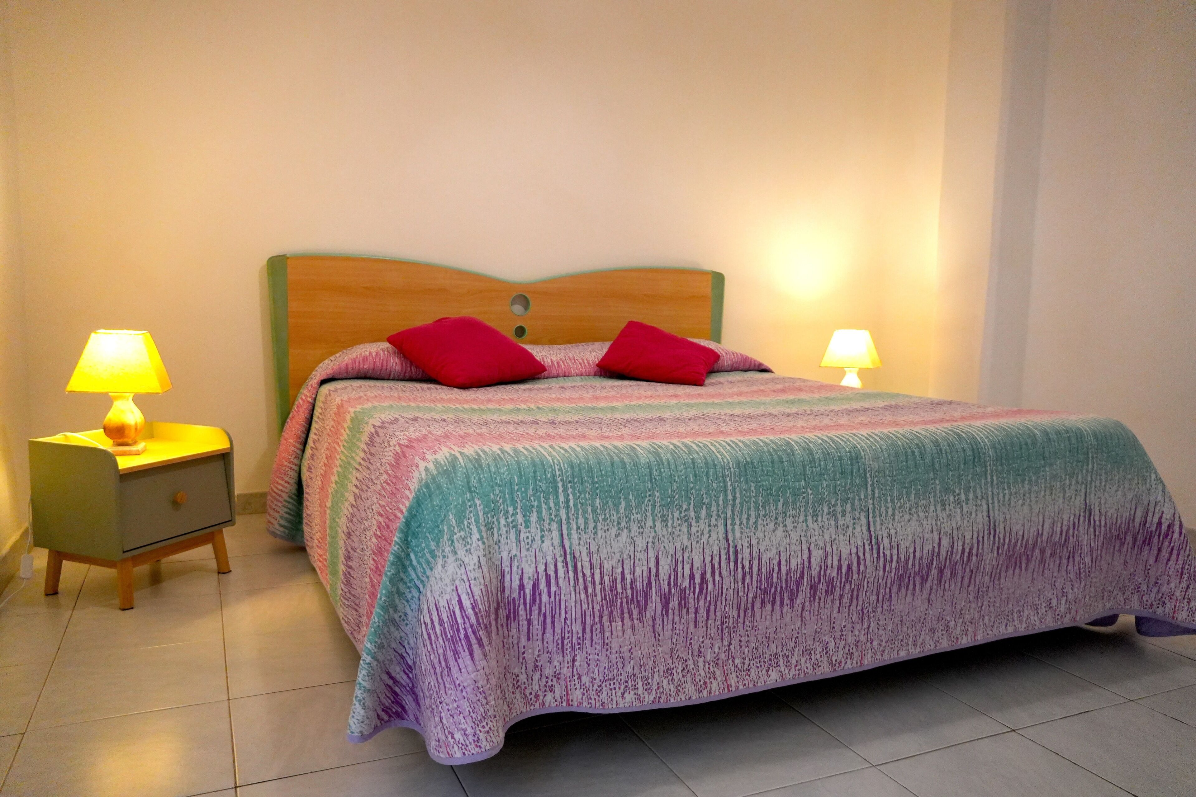 Uma cama com um cobertor colorido, dois travesseiros vermelhos e duas luminárias de cabeceira.