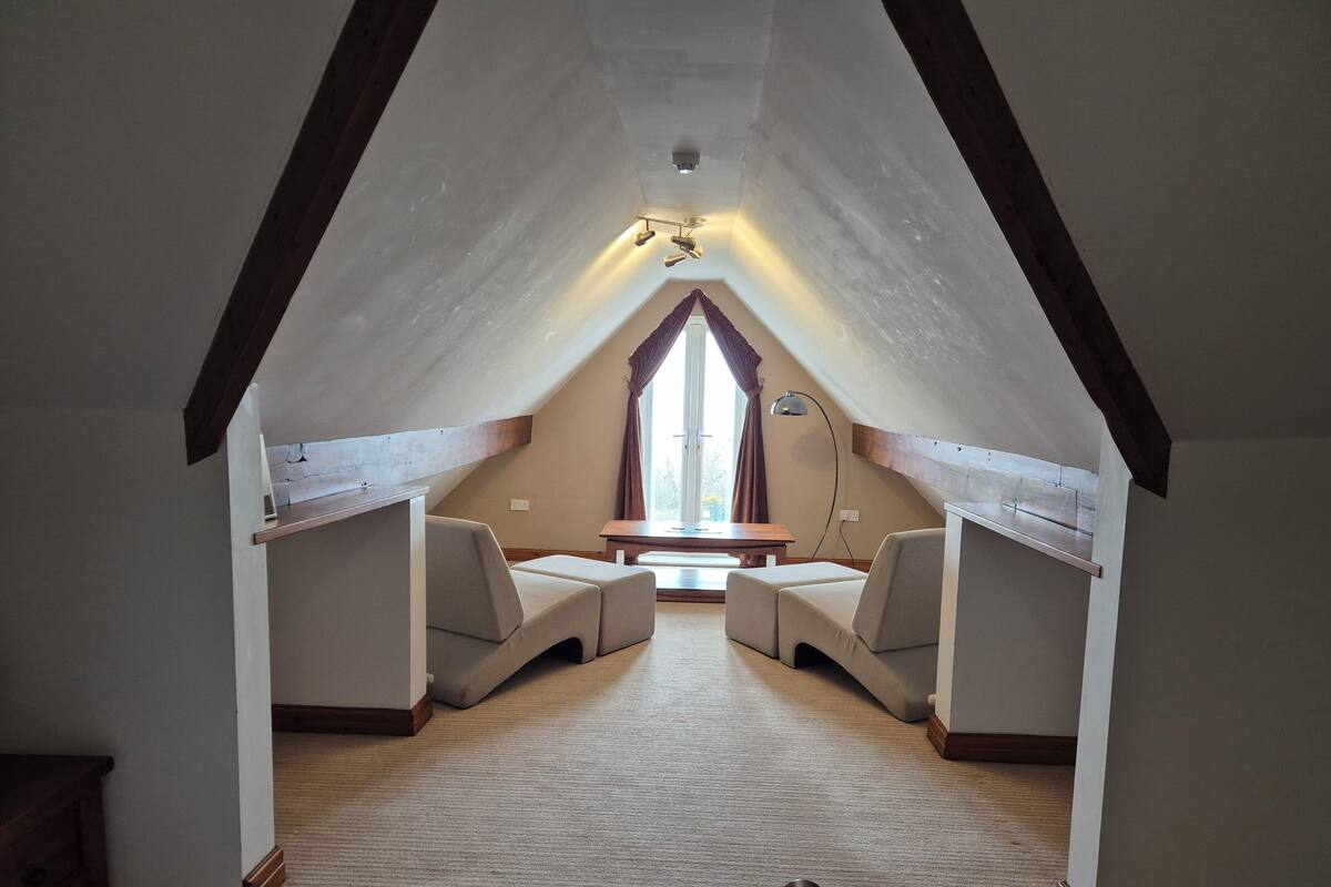 Quarto de sótão amplo, com pé direito alto, teto inclinado, uma janela grande com cortinas e um banco de madeira.