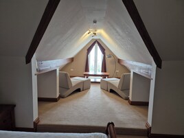 Quarto de sótão amplo, com pé direito alto, teto inclinado, uma janela grande com cortinas e um banco de madeira.