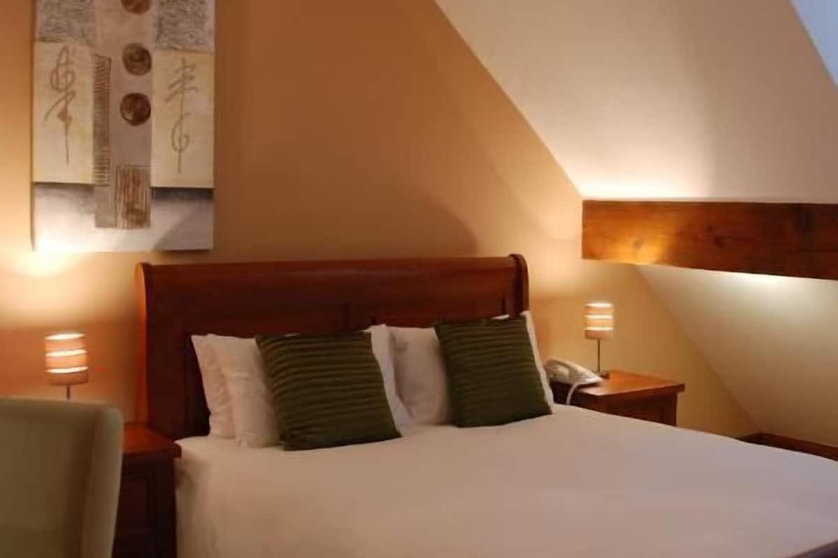 Quarto com cabeceira de madeira, cama com roupa de cama branca, duas mesas de cabeceira com luminárias e uma obra de arte na parede.