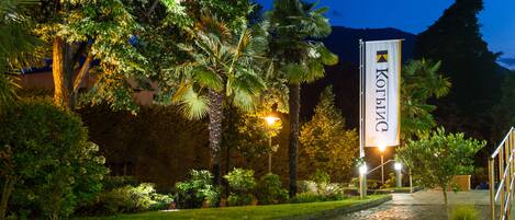 Vista noturna da entrada de um hotel com uma placa indicando 'HOTEL', cercada por vegetação exuberante e um caminho de pedras.
