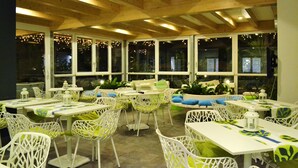Um espaço de refeições interno com mesas e cadeiras brancas, grandes janelas e luzes de Natal.