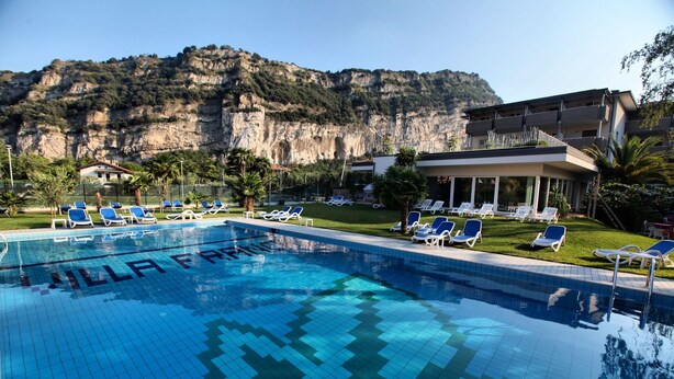 Piscina com espreguiçadeiras, montanhas ao fundo e um prédio com varandas.