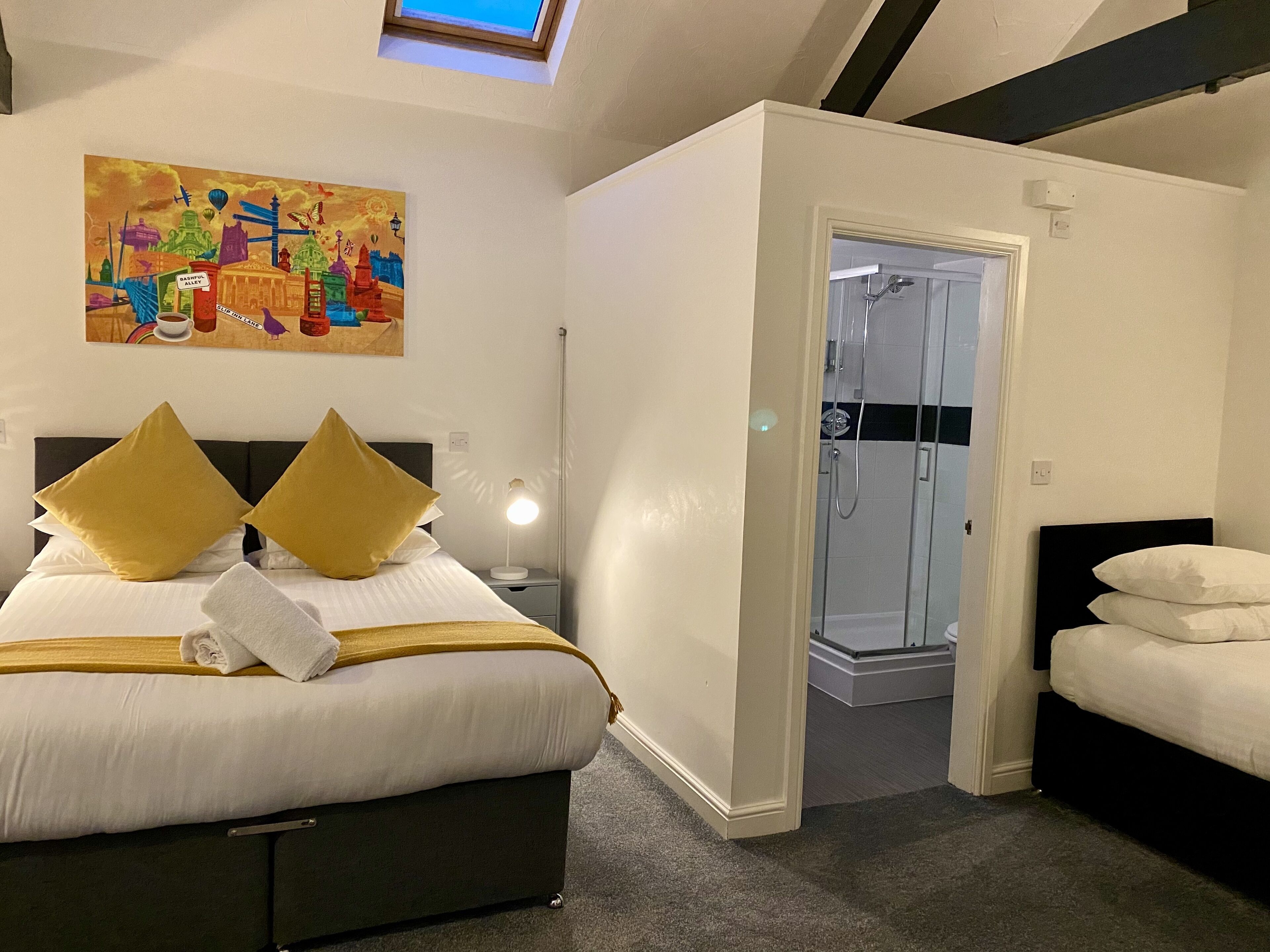 Quarto de hotel com cama, mesa de cabeceira, abajur, chuveiro e um quadro colorido na parede.