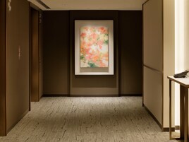 Um corredor com um quadro abstrato emoldurado na parede, uma porta à esquerda e uma pequena mesa com um dispositivo à direita.
