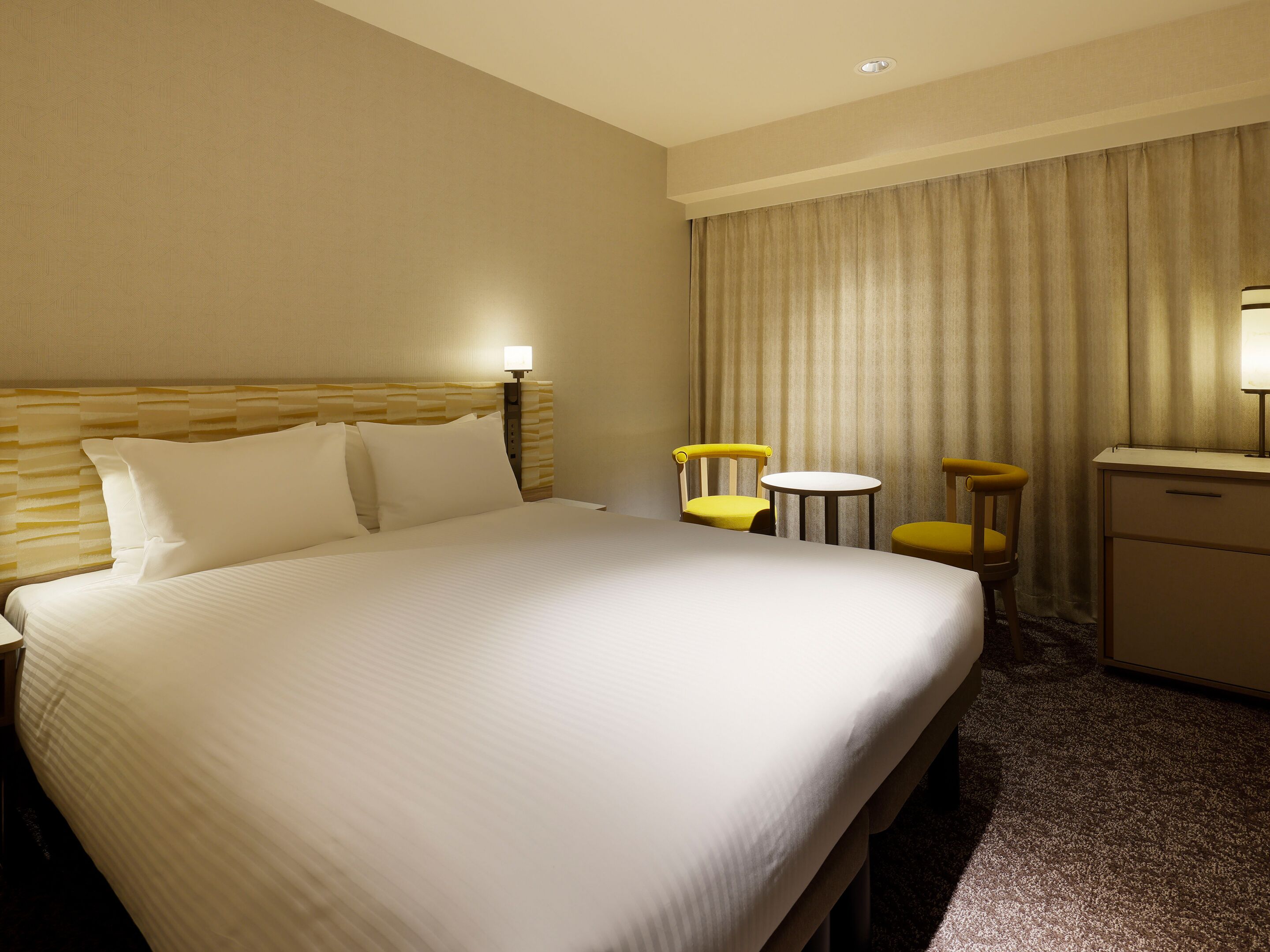 Quarto de hotel com uma cama grande, duas cadeiras, uma mesa pequena e uma escrivaninha.