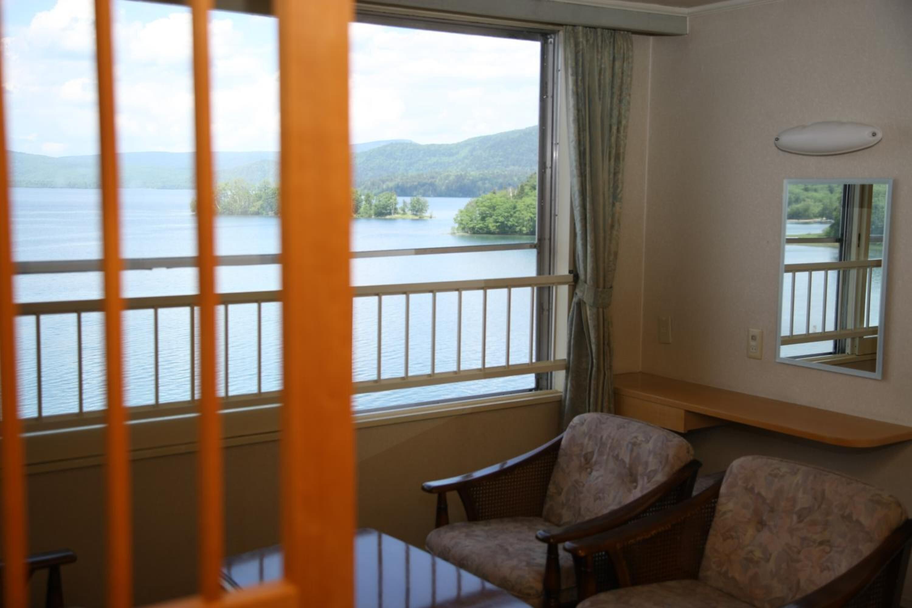 Quarto com vista para um lago e montanhas, uma cadeira, uma mesinha e um espelho na parede.