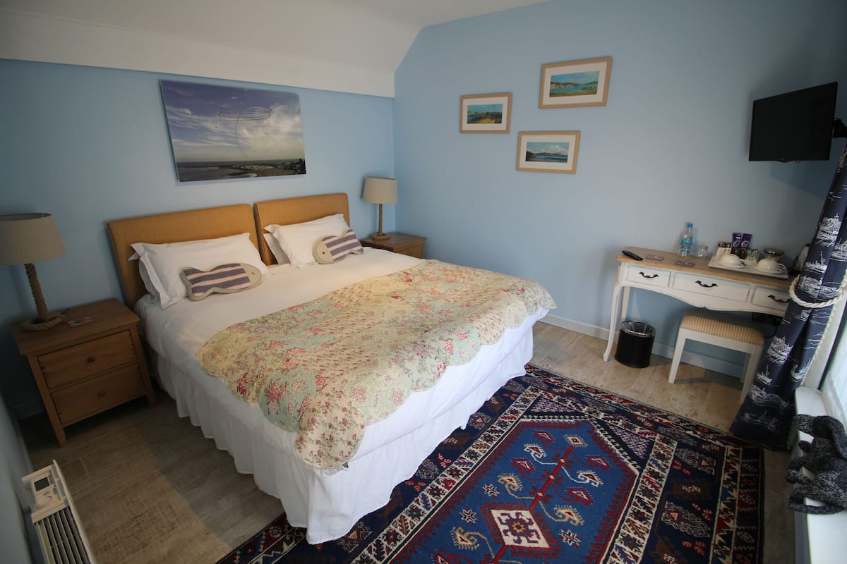 Quarto com cama, mesas de cabeceira, escrivaninha, cadeira e televisão.