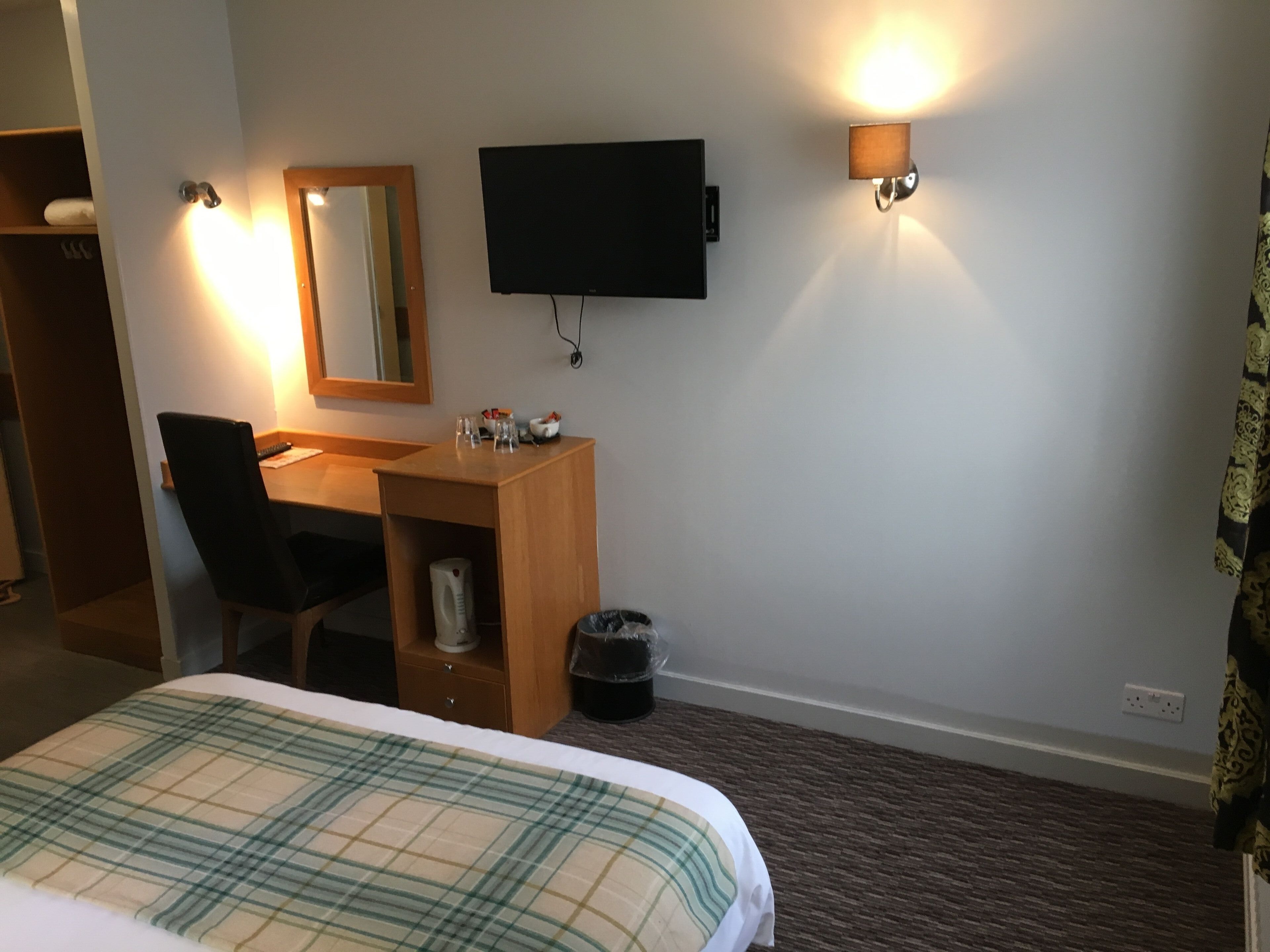 Quarto de hotel com cama, escrivaninha, cadeira, televisão e lixeira.