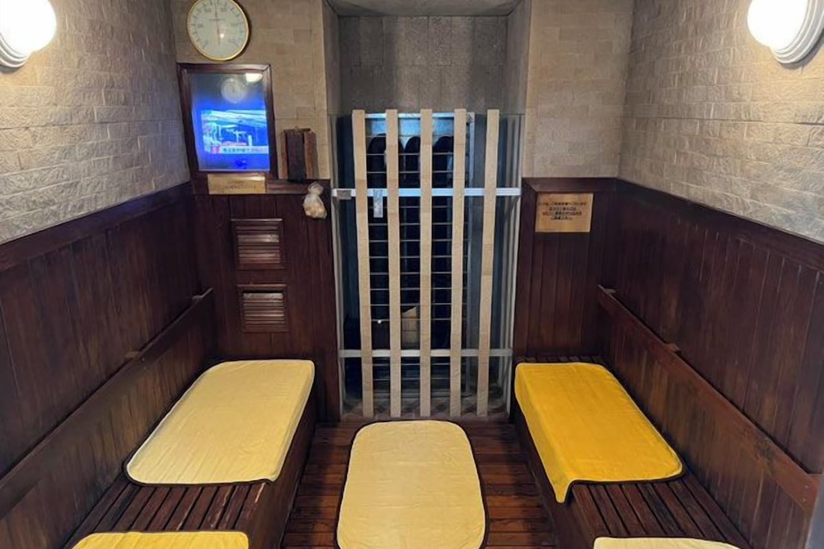 Um quarto pequeno e fechado, com paredes de madeira e chão de azulejos. Há três camas com roupa de cama amarela, um relógio na parede e uma televisão em uma prateleira.