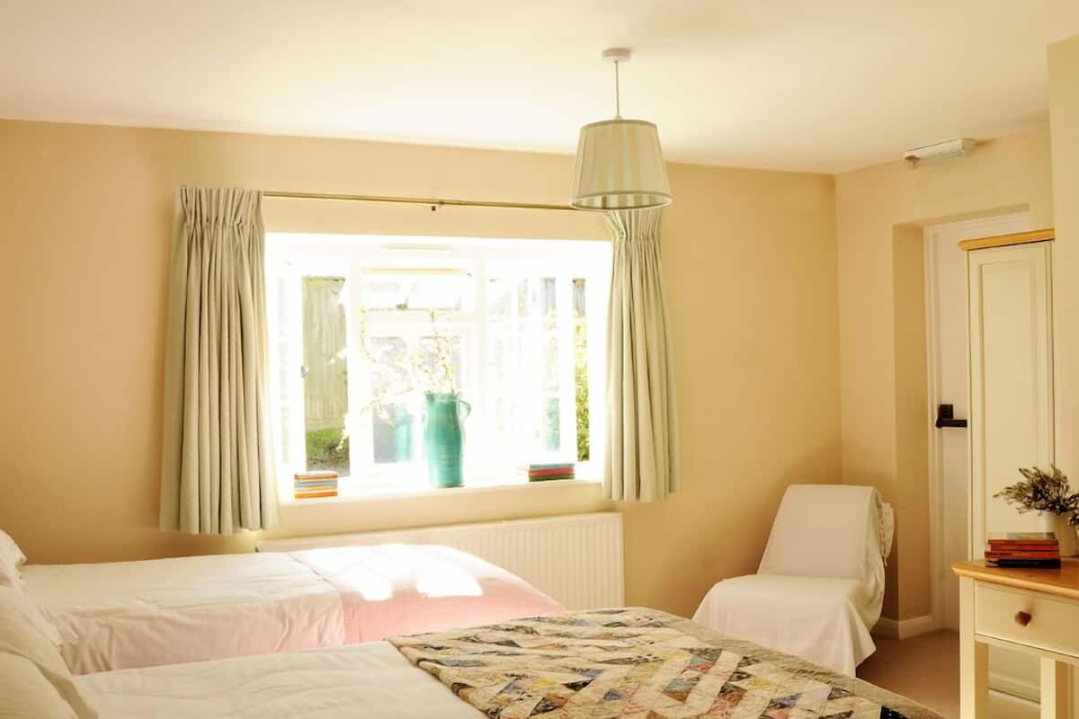 Quarto com duas camas, janela com cortinas, mesa de cabeceira com abajur e uma cadeira.