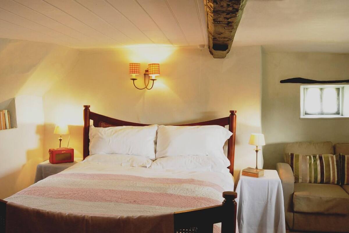 Quarto com cama de madeira, duas luminárias de apoio, uma mesinha com abajur, uma cadeira e uma janela com cortinas.