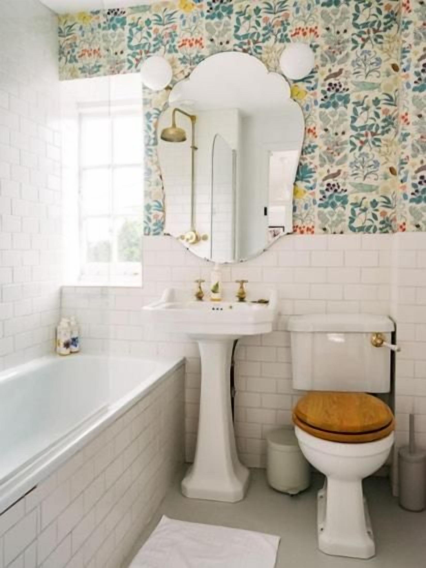 Uma casa de banho com uma banheira branca, uma pia assentada em pedestal, uma sanita com assento de madeira e um espelho com um padrão floral na parede.