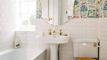 Uma casa de banho com uma banheira branca, uma pia assentada em pedestal, uma sanita com assento de madeira e um espelho com um padrão floral na parede.