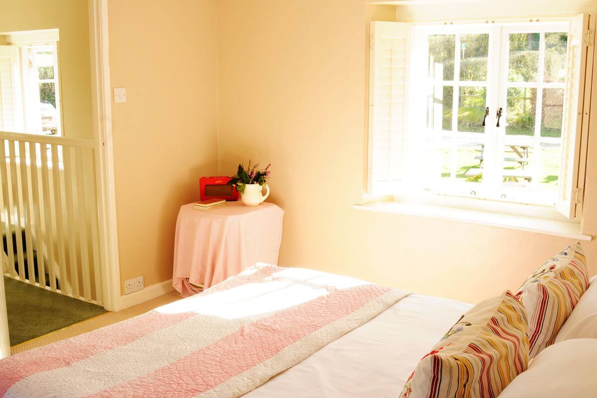 Quarto com cama, janela com vista para o verde, pequena mesa com rádio e um vaso de flores, e um corrimão branco.