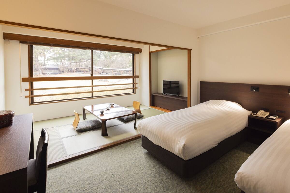 Quarto de hotel com uma cama, uma escrivaninha com cadeira, televisão e uma janela com persianas.