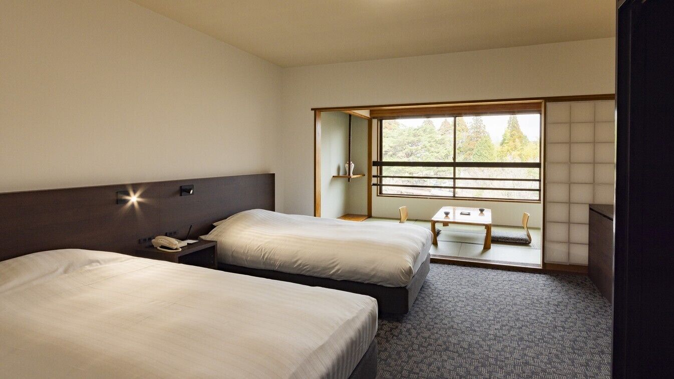 Quarto de hotel com duas camas, uma mesinha, uma mesaninha de cabeceira, uma luminária e uma janela com vista para árvores.