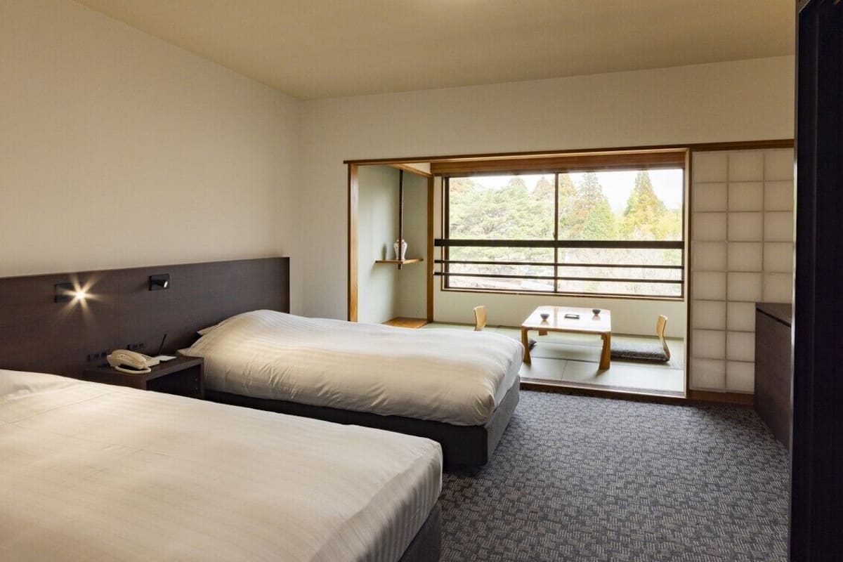 Quarto de hotel com duas camas, uma mesinha, uma mesaninha de cabeceira, uma luminária e uma janela com vista para árvores.