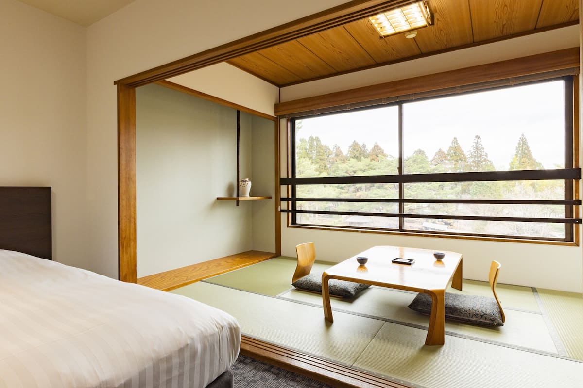 Um quarto em estilo japonês tradicional, com piso de tatame, cama, mesa baixa com almofadas e uma grande janela com vista para árvores.